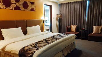 Quarto de hotel com uma cama grande, uma cadeira, uma mesinha e vista para o exterior pela janela.