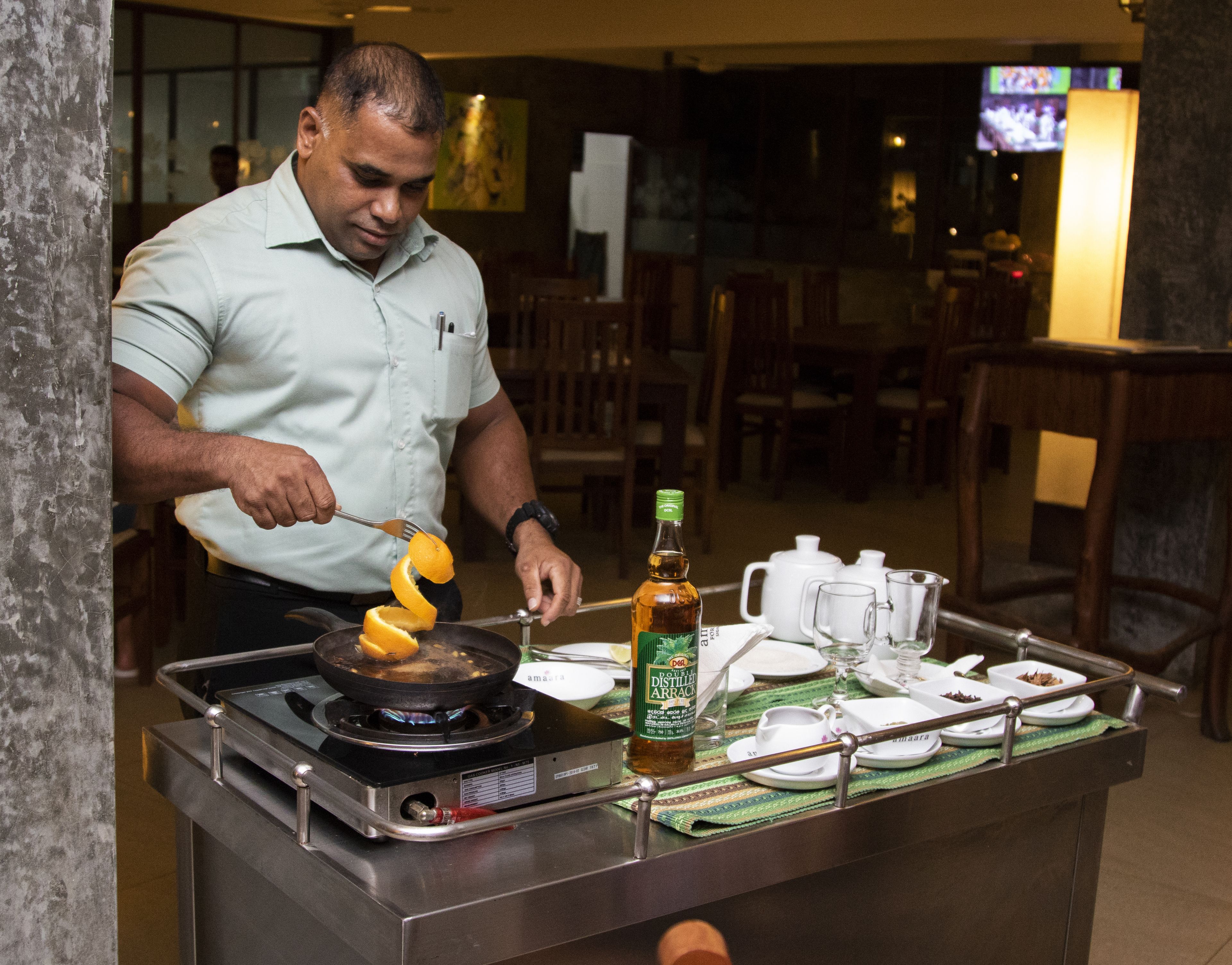 Um homem cozinha em um fogão portátil em um restaurante, com uma mesa posta para refeição, incluindo pratos, copos e uma garrafa de bebida alcoólica.