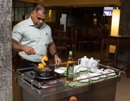 Um homem cozinha em um fogão portátil em um restaurante, com uma mesa posta para refeição, incluindo pratos, copos e uma garrafa de bebida alcoólica.