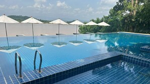 Uma piscina infinita azul cristalina, com borda em azulejos, rodeada por guarda-sóis brancos e árvores.