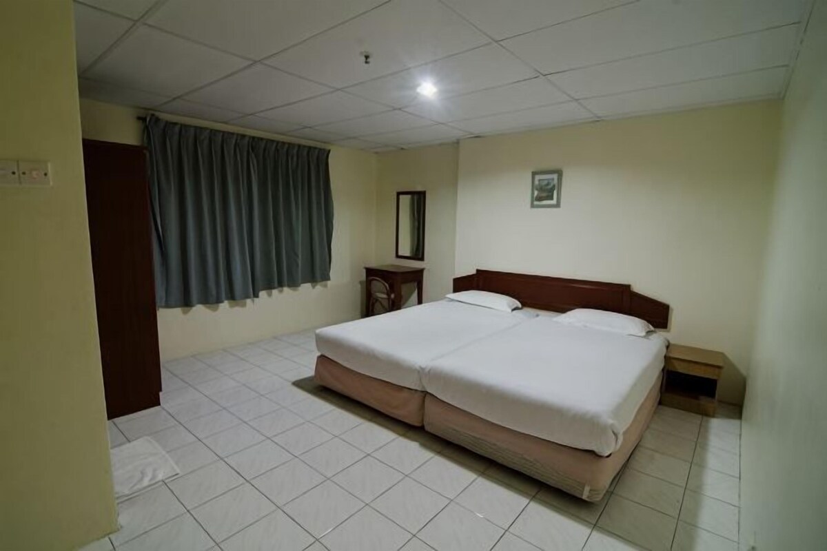 Uma cama de casal, uma mesinha de cabeceira pequena, um espelho, uma janela com cortinas e um piso ladrilhado.
