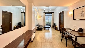 Quarto de hotel moderno com piso de madeira, um sofá, uma mesa de jantar com cadeiras, um espelho grande e vista para a cidade pela janela.