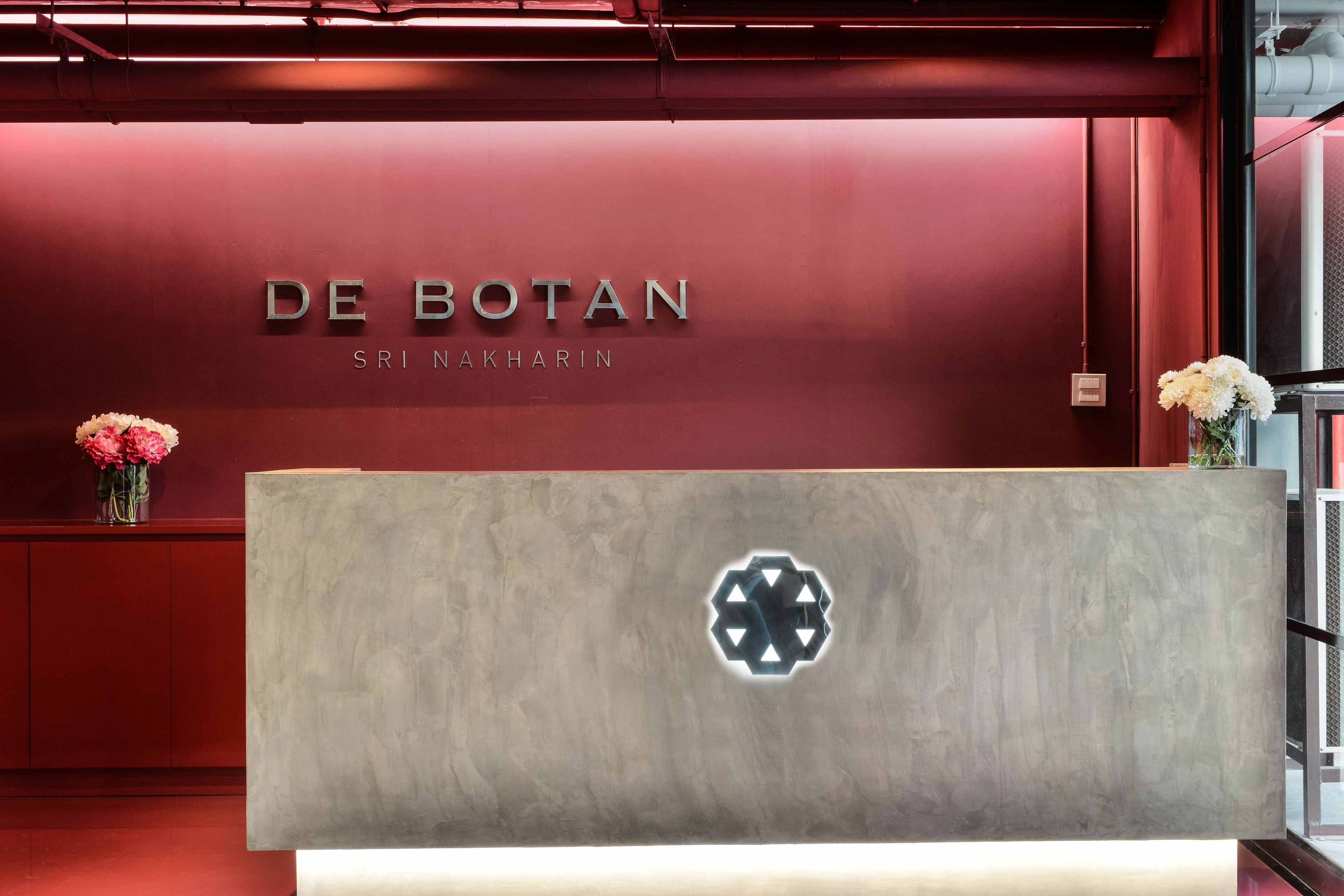 Uma mesa de recepção com um logotipo, uma parede vermelha com o nome 'DE BOTAN SRI NARHARI' acima dela, e um vaso com flores à esquerda.