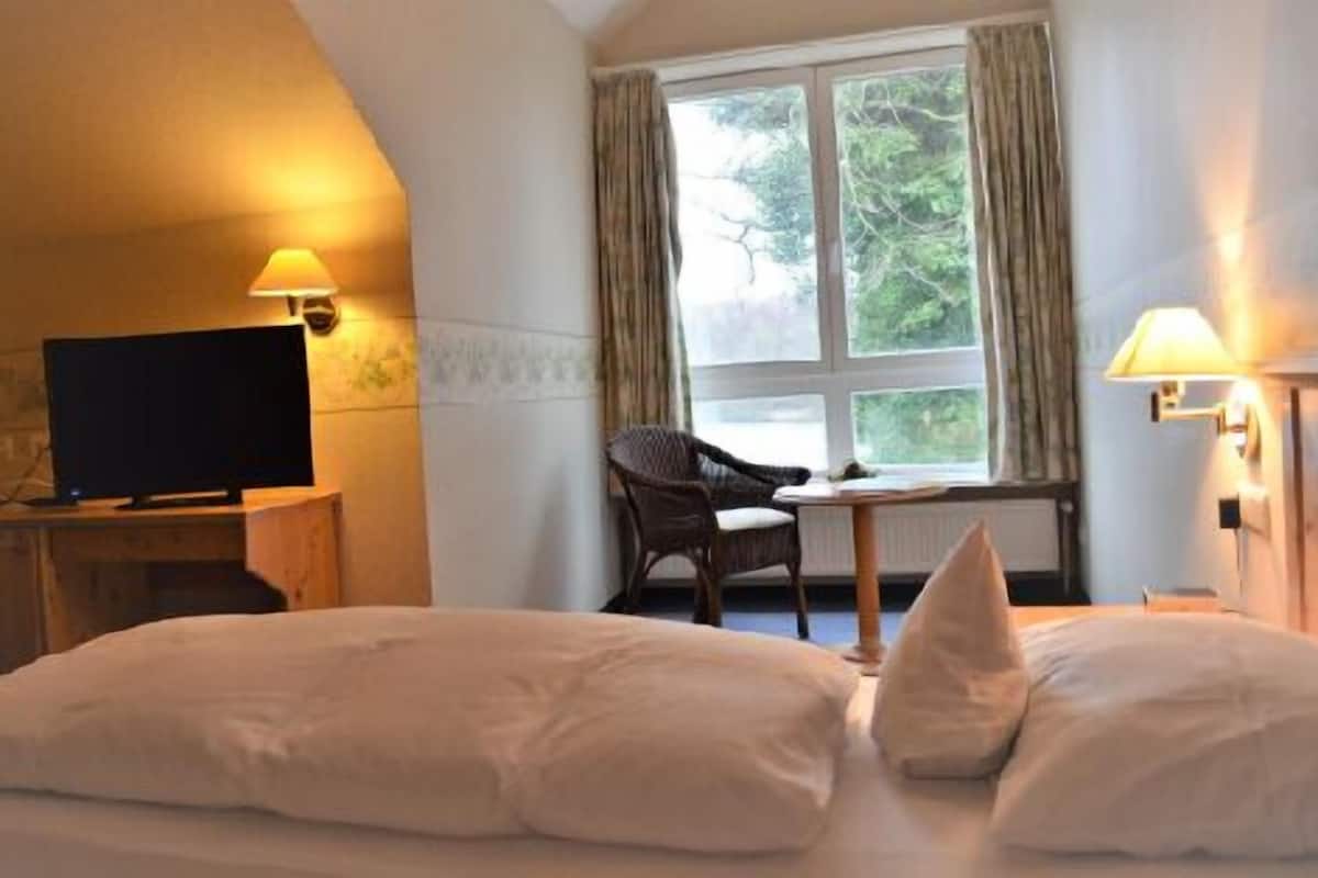 Quarto de hotel com cama, televisão, cadeira e janela com cortinas.