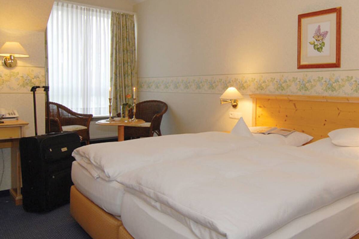 Quarto de hotel com uma cama grande, uma escrivaninha com abajur, uma cadeira e uma mesinha de centro com um vaso.