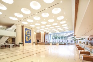 Um hall amplo com design moderno, apresentando um grande balcão de recepção, uma escadaria e um janelão com vista para o exterior.