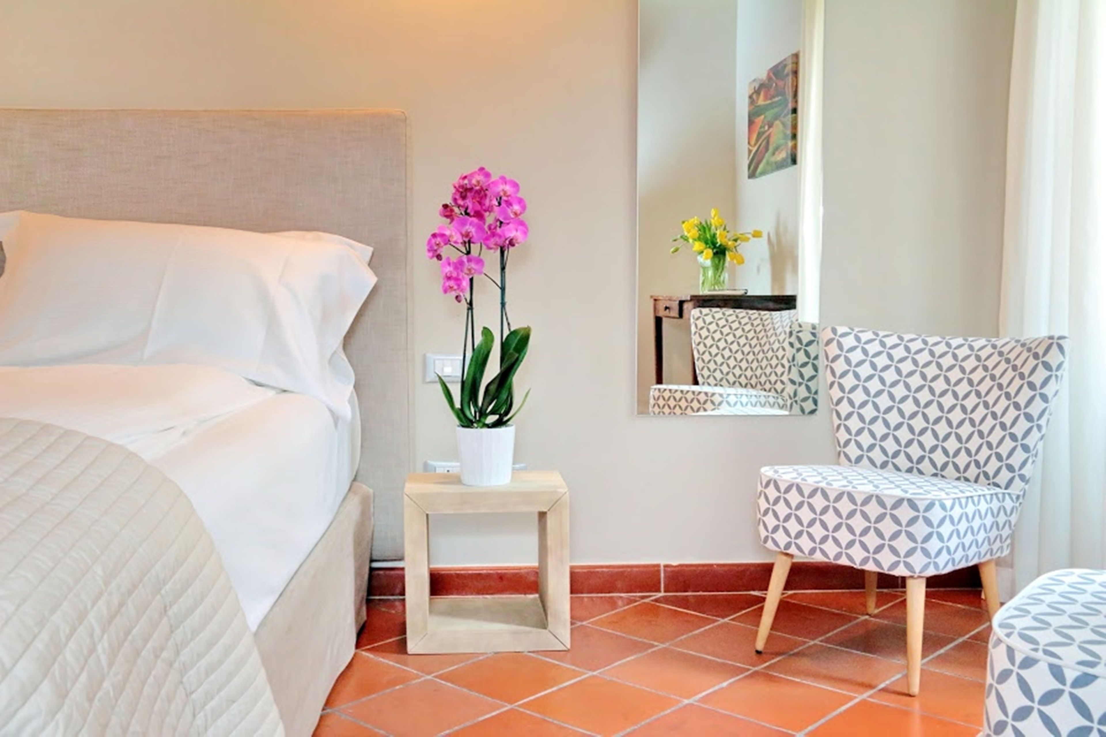 Quarto de hotel com uma cama, uma pequena mesa com uma planta em vaso, uma cadeira e um espelho na parede.