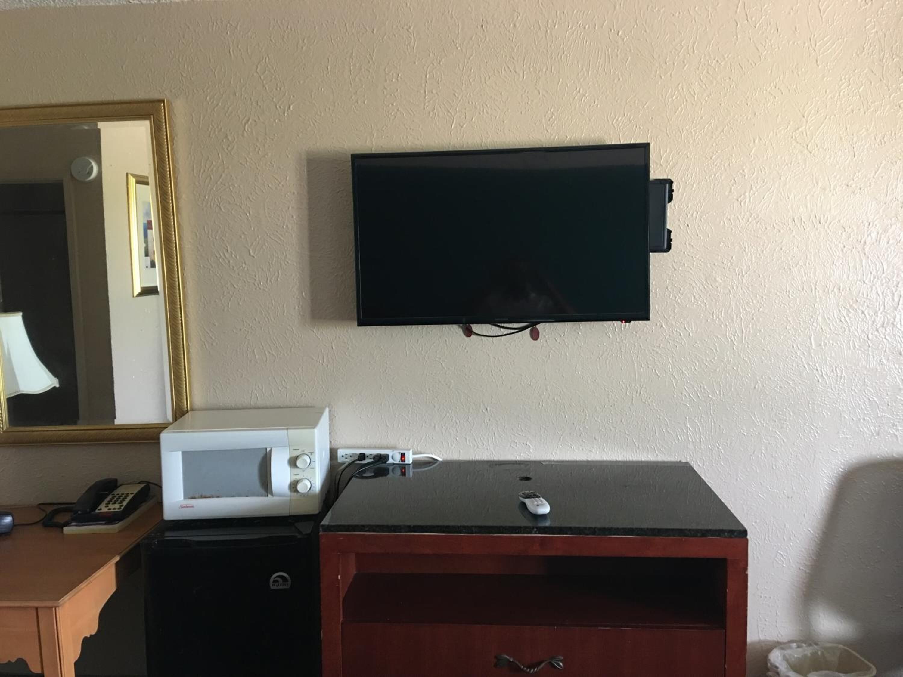 Quarto de hotel com televisão na parede, micro-ondas, telefone e um pequeno armário com gaveta.