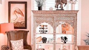 Um quarto com uma estante decorativa repleta de objetos diversos, uma cadeira de vime, um sofá estofado e uma obra de arte emoldurada na parede.