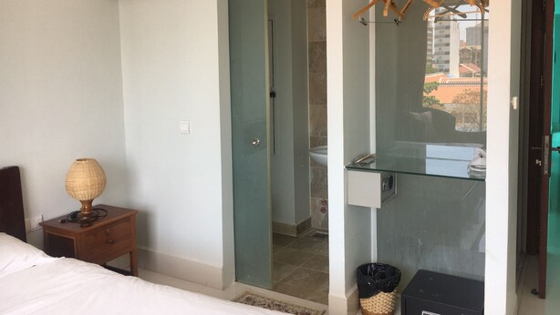 Quarto de hotel com cama, mesa de cabeceira com abajur, ar condicionado, guarda-roupa com espelho e vista para o exterior pela janela.