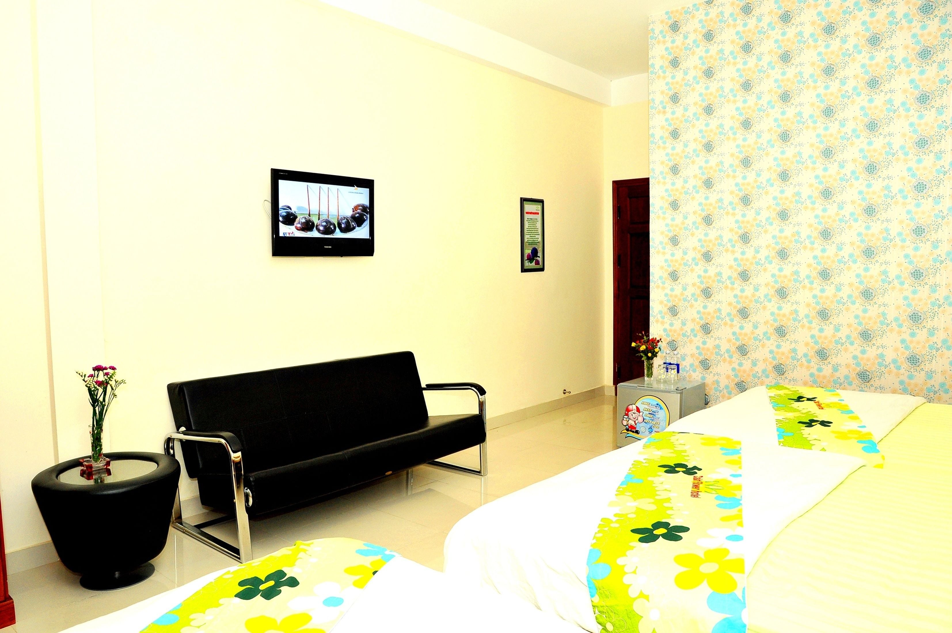 Quarto de hotel com cama, um sofá preto, uma mesa pequena com um vaso de flores, televisão na parede e cortina estampada.
