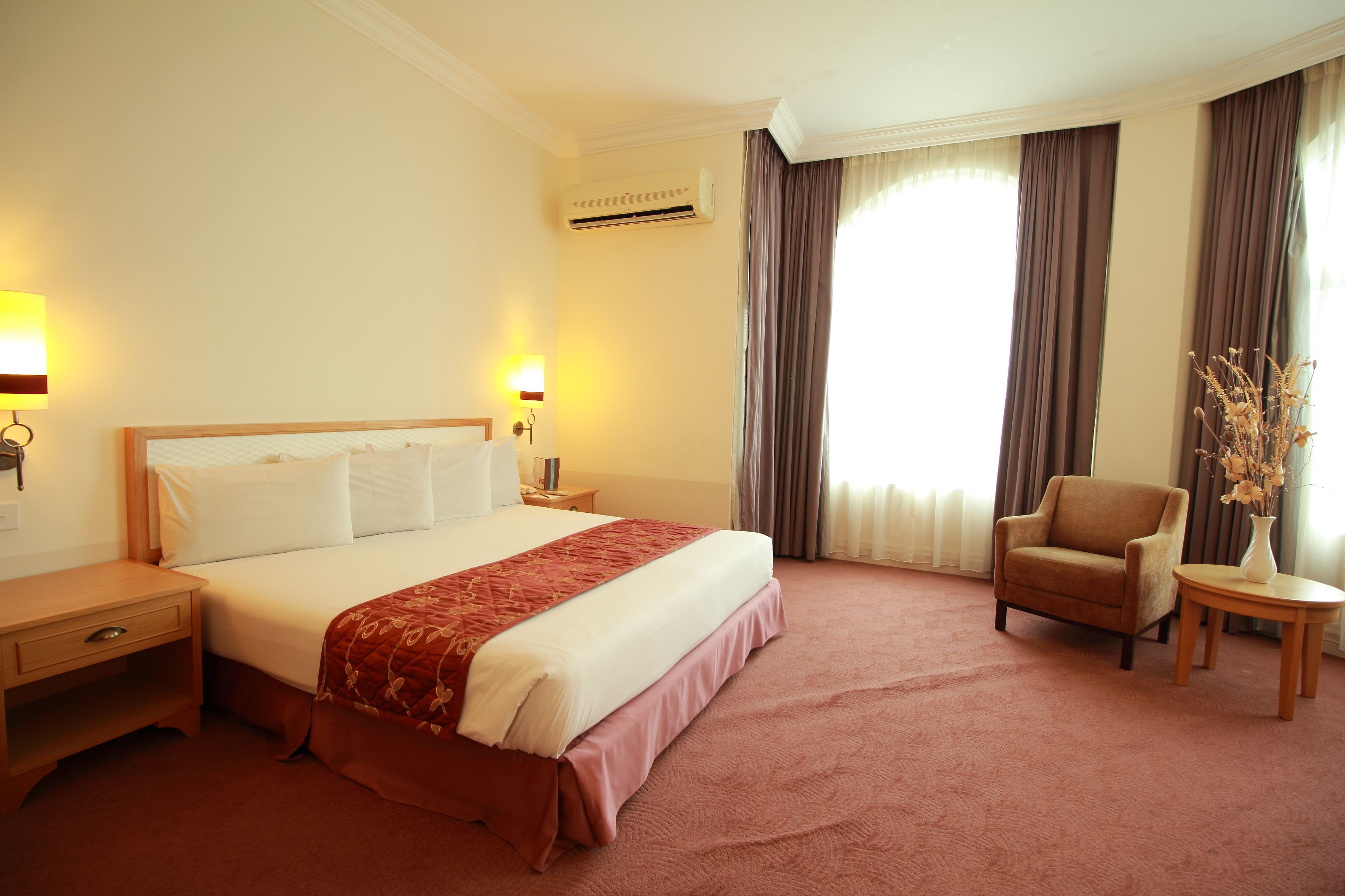 Quarto de hotel com uma cama grande, uma cadeira, uma mesinha e uma luminária.