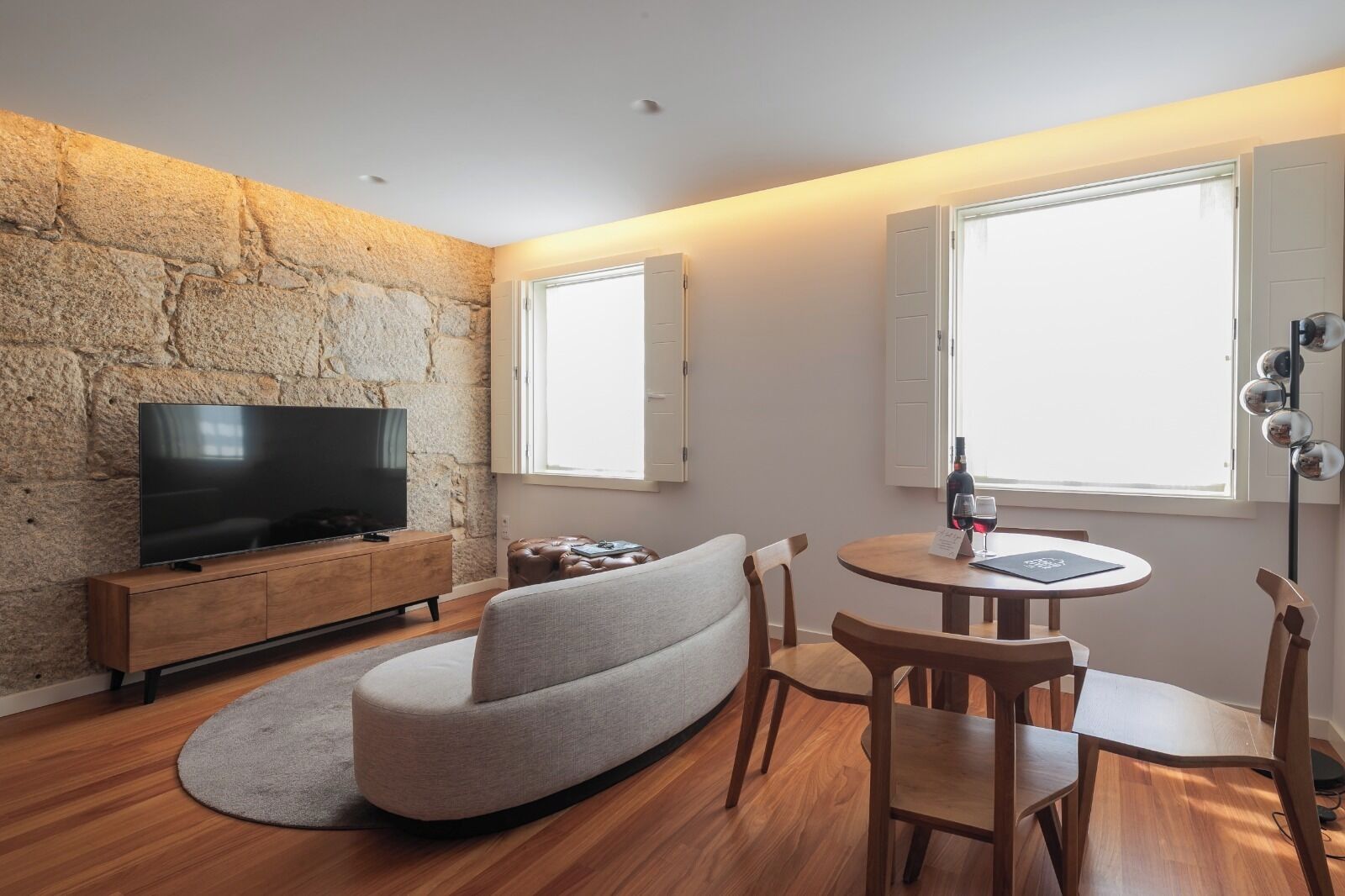 Uma sala de estar moderna com parede de pedra, televisão de tela plana, sofá cinza, piso de madeira, mesa de jantar redonda com cadeiras e uma janela com persianas brancas.