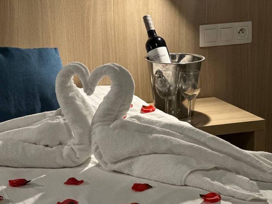 Cama com toalha branca disposta em formato de cisnes, garrafa de vinho, copos em recipiente de metal e pétalas de rosa espalhadas sobre a cama.