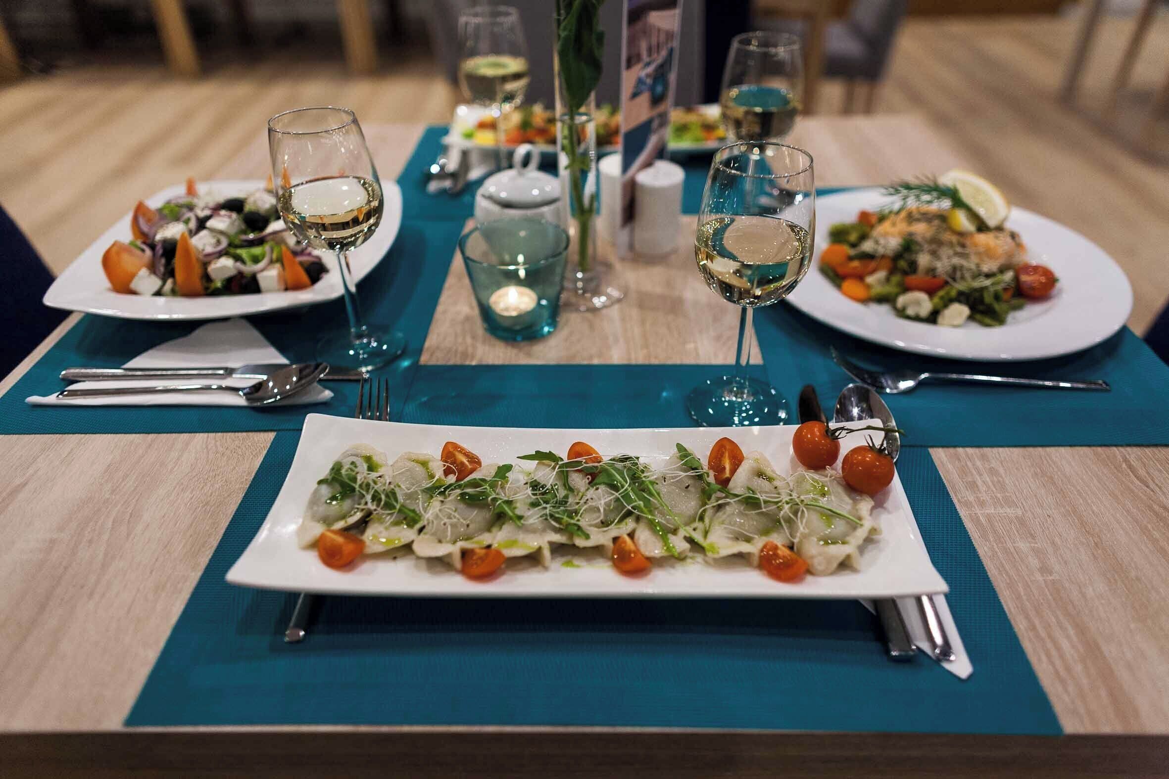 Mesa de jantar posta com pratos de comida, copos de vinho branco e um jogo americano azul-turquesa.