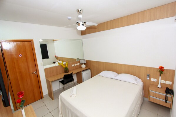 Quarto de hotel com cama, secretária com cadeira, pequeno frigorífico, mesa de cabeceira com telefone e espelho sobre a secretária.