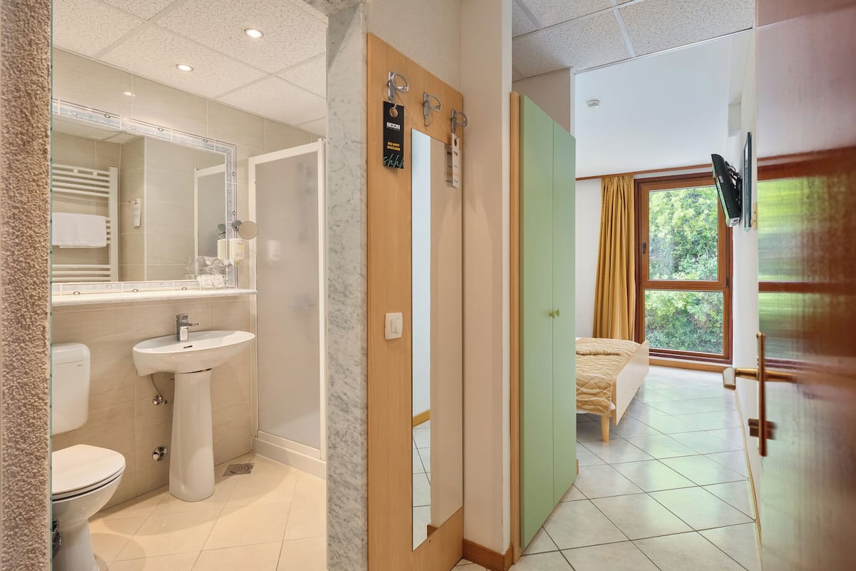 Uma casa de banho com lavatório, sanita e espelho, um quarto com cama e janela, e um corredor com porta.