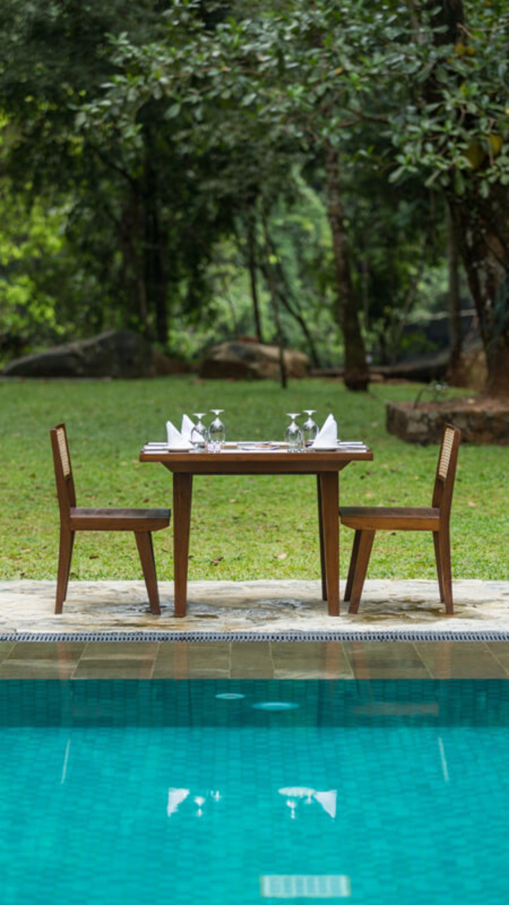 Uma área de refeições à beira da piscina, com uma mesa e duas cadeiras, rodeada por um jardim verdejante.