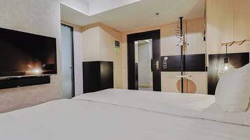 Quarto de hotel com televisão de tela plana fixada na parede, cama com roupa branca, armário de madeira e luminária moderna.