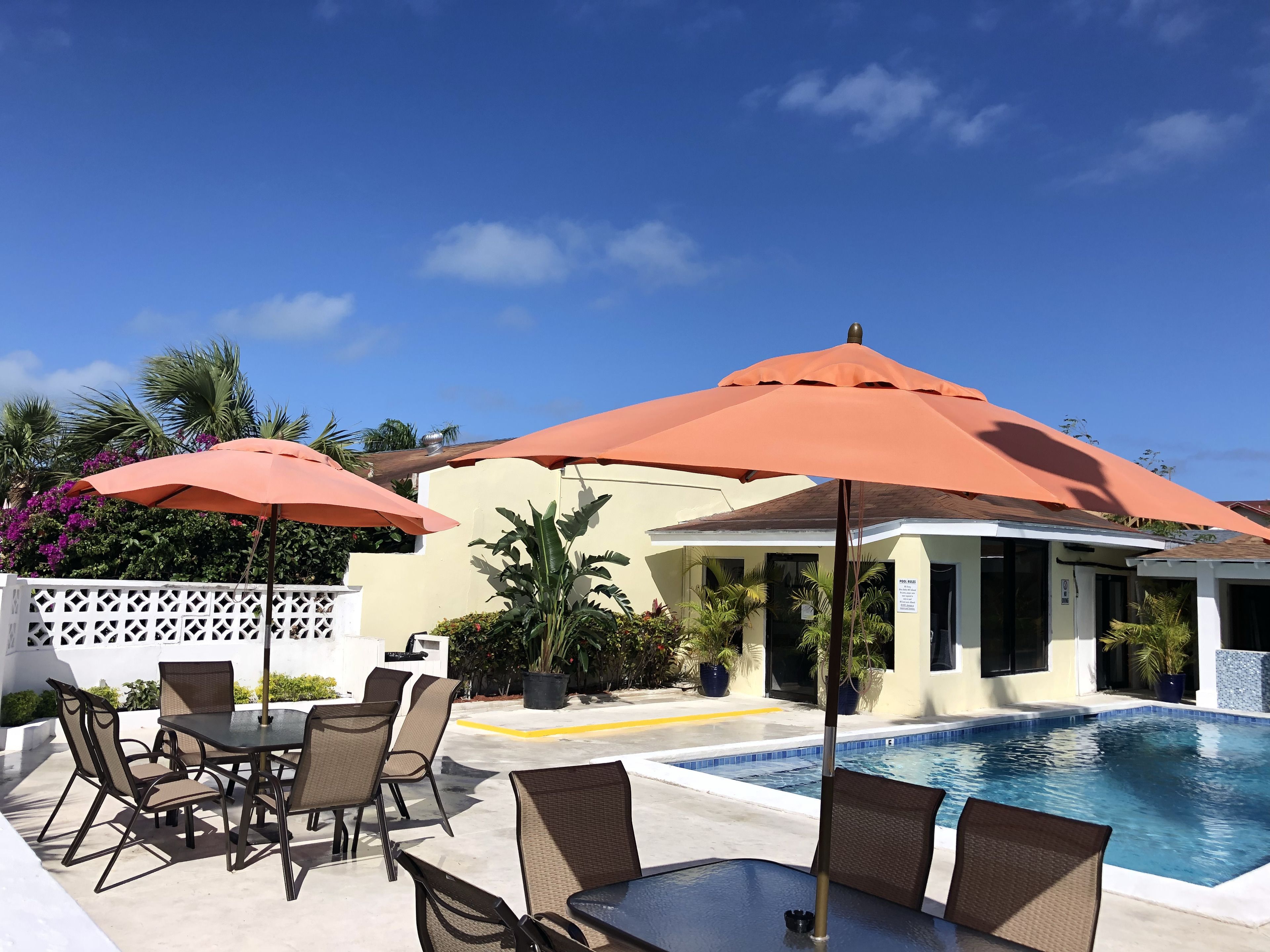 Uma área de piscina externa com mesa e cadeiras, cercada por guarda-sóis e plantas em vasos.