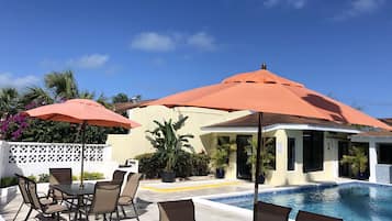 Uma área de piscina externa com mesa e cadeiras, cercada por guarda-sóis e plantas em vasos.
