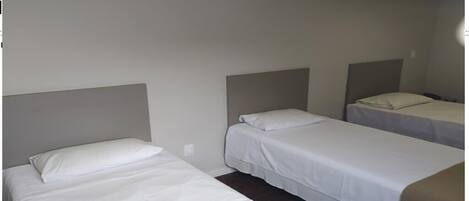 Quarto com três camas de solteiro alinhadas na parede, cada uma com cabeceira e roupa de cama branca.