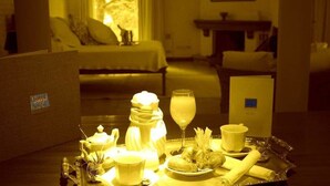 Uma bandeja com uma vela, um copo de bebida, uma xícara e doces sobre uma mesa em um quarto iluminado com uma cama e um criado-mudo ao fundo.