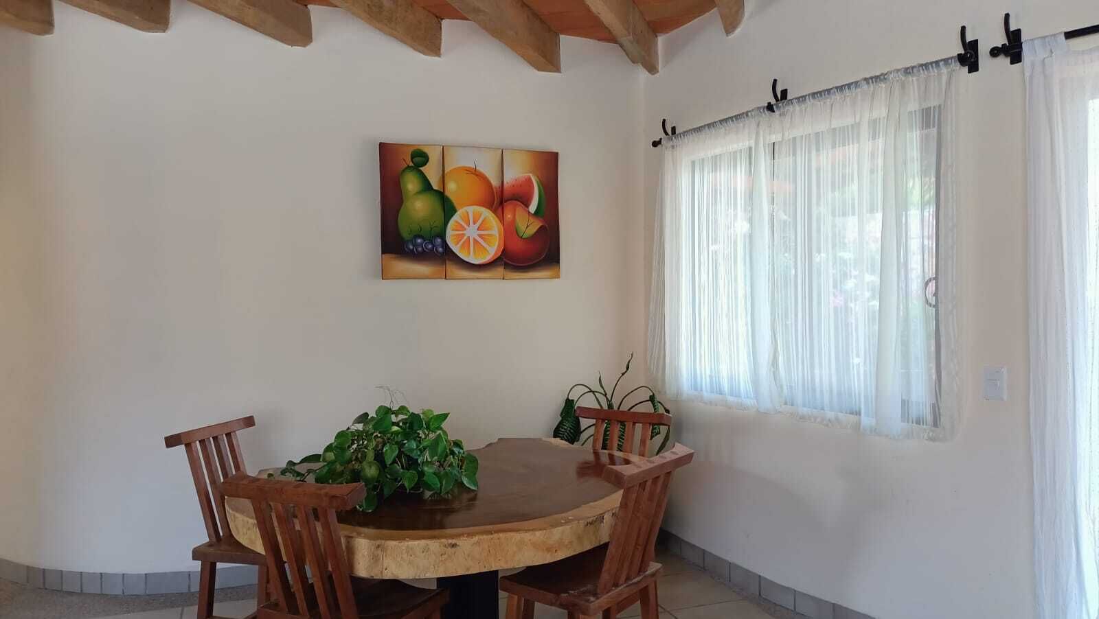 Uma área de refeições com mesa e cadeiras de madeira, um quadro de frutas na parede e uma janela com cortinas leves.