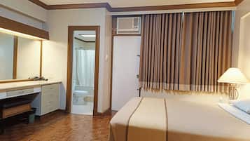 Quarto de hotel com cama, escrivaninha com gavetas, espelho, aparelho de ar condicionado e janela com cortinas.