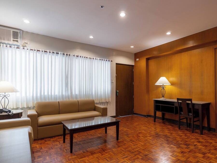 Quarto de hotel com sofá, mesa, abajur, escrivaninha com cadeiras e guarda-roupa de madeira.