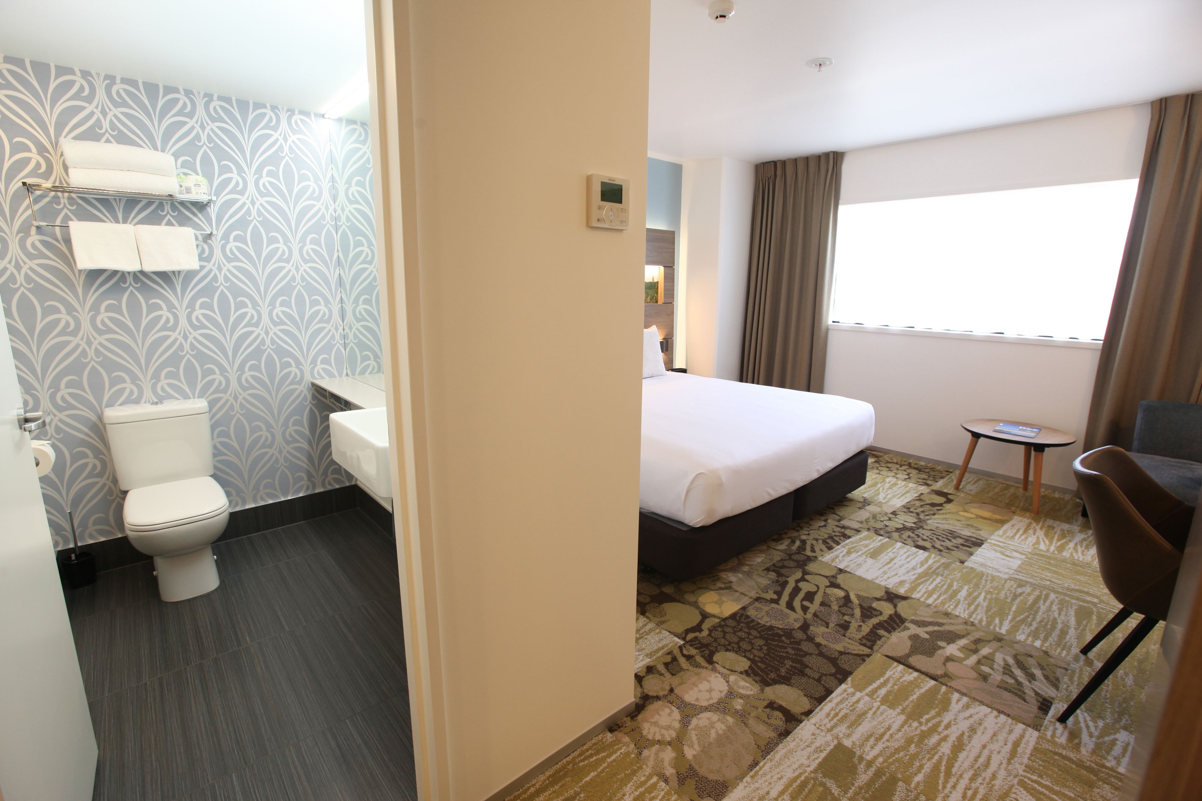 Quarto de hotel com cama, banheiro com vaso sanitário e pia, uma mesinha e uma cadeira.