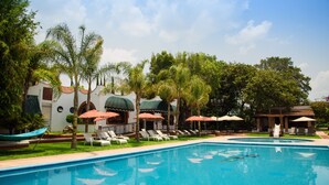 Piscina com espreguiçadeiras e guarda-sóis, cercada por palmeiras e um edifício com telhado em azulejos.