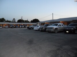Um estacionamento com vários veículos, incluindo uma caminhonete branca e um SUV preto, em frente a um edifício com telhado plano.