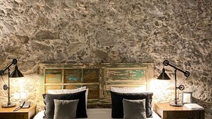 Quarto com parede de pedra, cabeceira de madeira, duas mesas de cabeceira com luminárias e uma cama com almofadas.