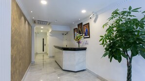 Uma área de recepção com piso de mármore, balcão de atendimento, retratos emoldurados na parede e um vaso de plantas.