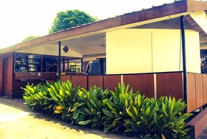 Um edifício com área externa coberta, um balcão com produtos expostos e uma fileira de plantas na frente.