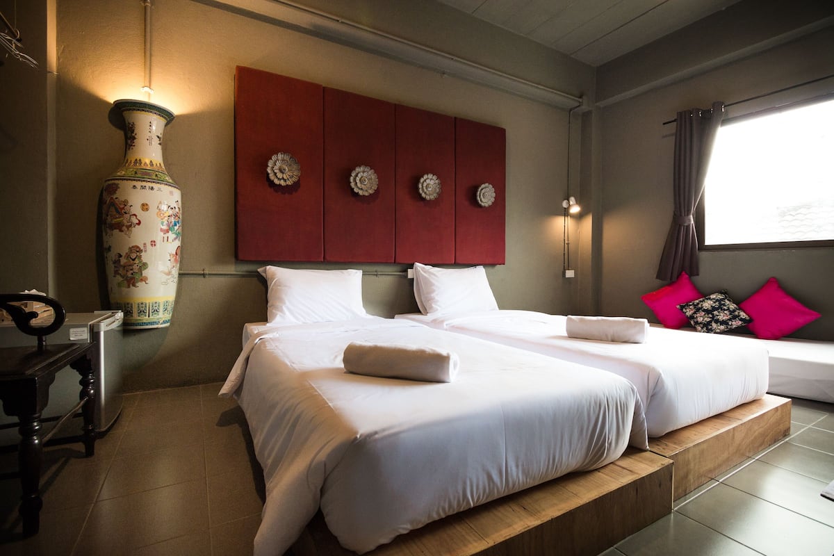 Quarto de hotel com duas camas, um vaso decorativo grande, um armário vermelho com pratos decorativos e uma janela com cortinas.