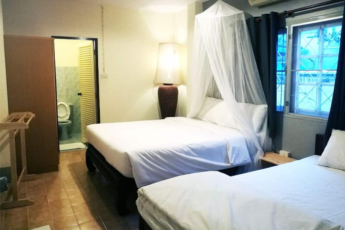 Quarto de hotel com duas camas, uma mesinha de cabeceira, uma luminária, uma janela com cortinas e uma porta para o banheiro.