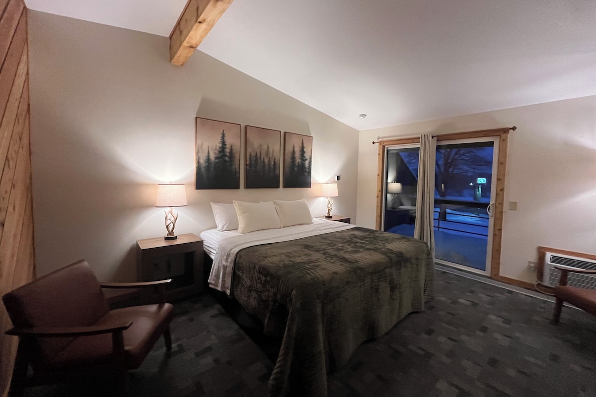 Quarto com cama, duas mesas de cabeceira com luminárias, uma cadeira e uma janela grande com vista para o exterior.