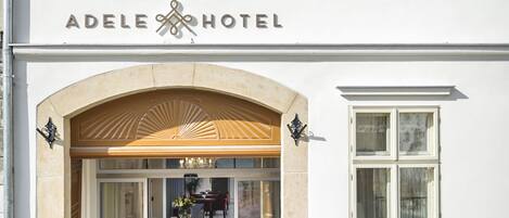 Um hotel de dois andares com fachada clássica, entrada central com claraboia e uma placa com o texto 'ADELE HOTEL'.