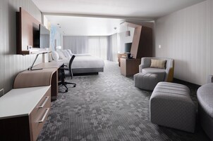 Quarto de hotel moderno com cama, escrivaninha, poltrona, televisão e um espelho grande.