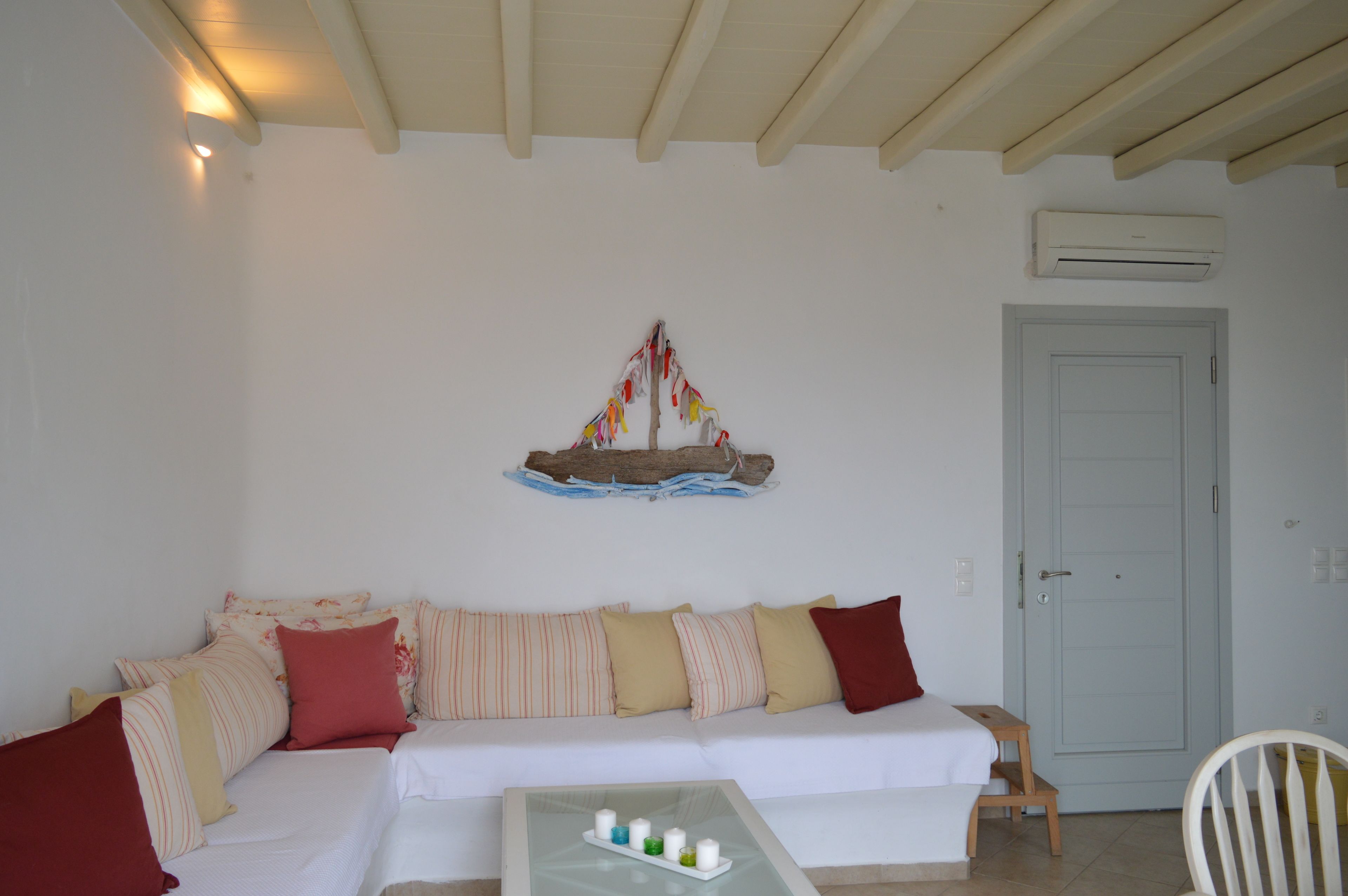 Sala de estar com sofá branco, almofadas vermelhas e bege, mesa de centro de vidro com velas, cadeira de madeira, porta e decoração na parede.