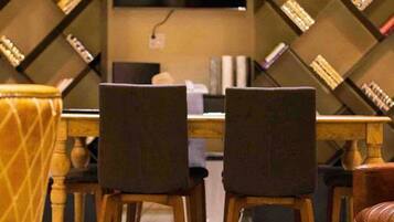 Uma área de refeições com mesa e cadeiras de madeira, uma televisão de tela plana na parede e prateleiras com diversos objetos.