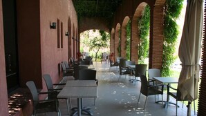 Um café ao ar livre com mesas e cadeiras, coberto por um telhado com arcos e vegetação nas paredes.