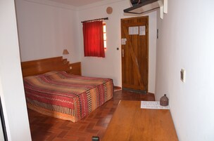 Quarto com cama, porta de madeira, cortina vermelha, abajur e mesinha.