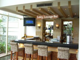 Um bar lounge no lobby com uma TV na parede, balcão com banquetas e diversas garrafas e copos nas prateleiras.