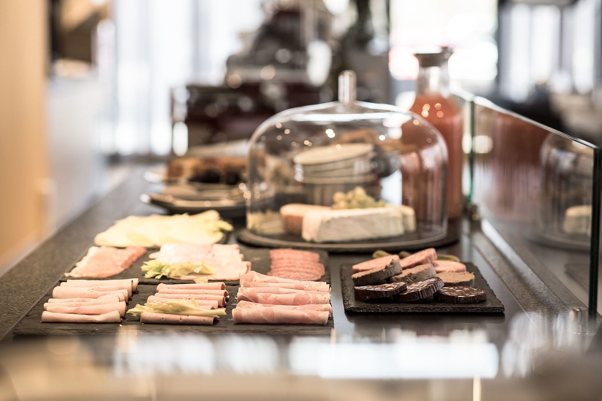 Buffet com diversos tipos de carne e queijo, incluindo fatias de presunto e queijo, e uma cúpula de vidro sobre uma seleção de queijos.