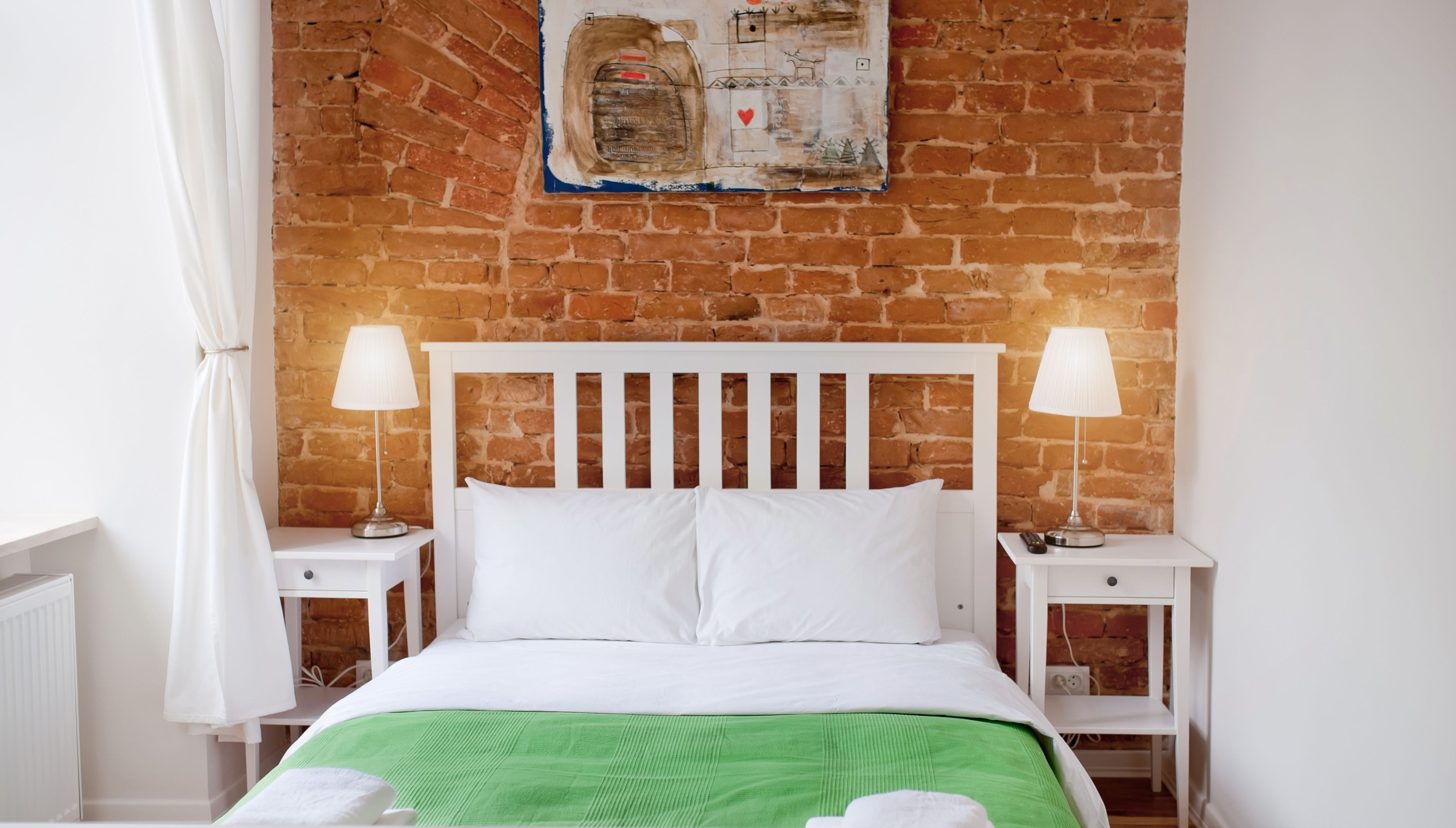 Uma cama com roupa de cama branca, duas mesas de cabeceira com abajures, uma parede de tijolos e um quadro sobre a cama.