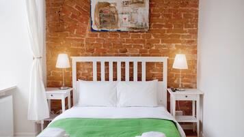Uma cama com roupa de cama branca, duas mesas de cabeceira com abajures, uma parede de tijolos e um quadro sobre a cama.
