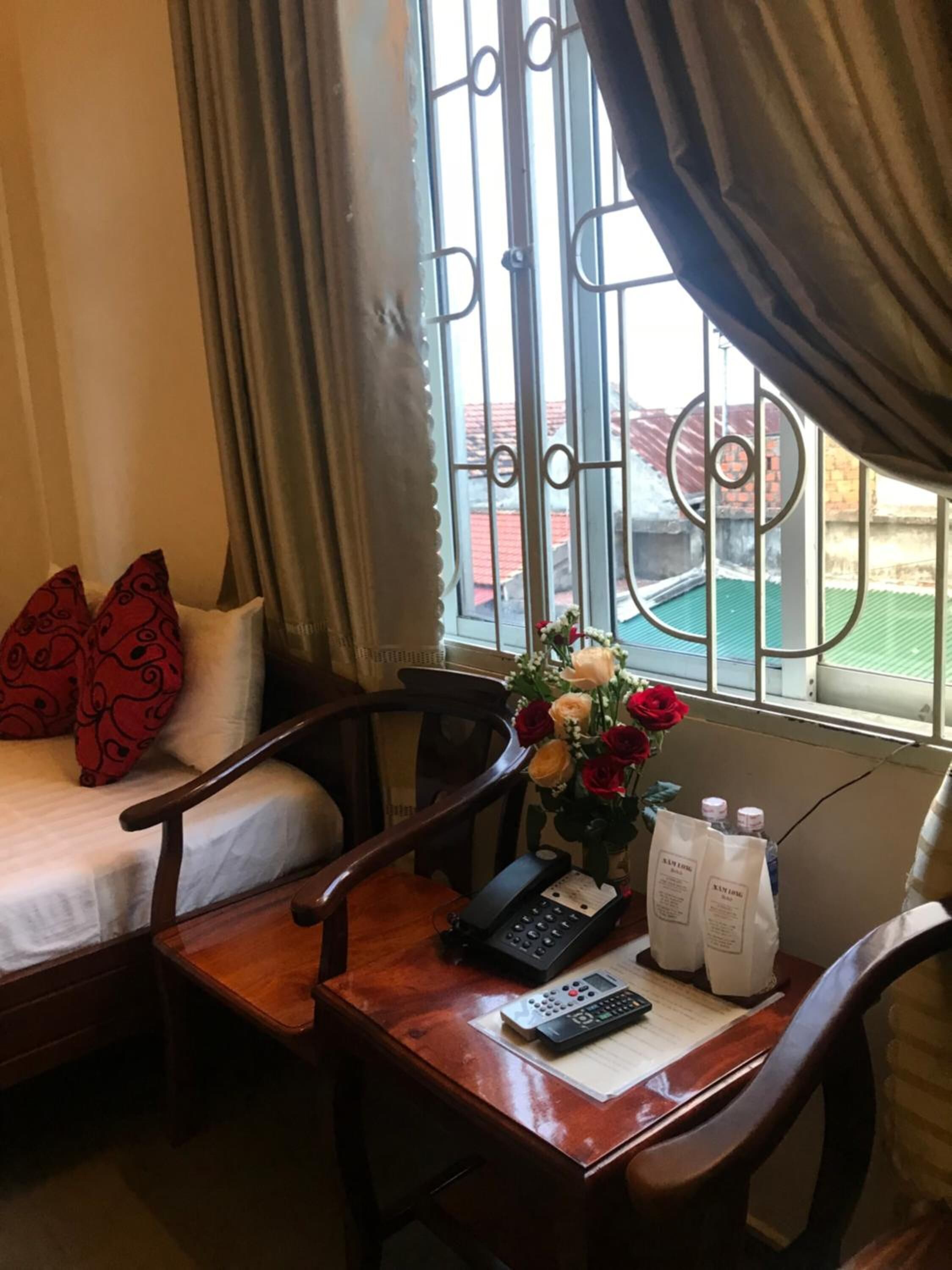 Quarto de hotel com uma cama, uma escrivaninha com telefone e um vaso de flores, e uma janela com cortinas.